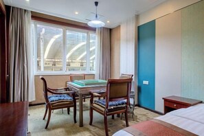 Quarto de hotel com uma cama, uma escrivaninha com cadeiras, uma janela com cortinas e vista para um edifício.