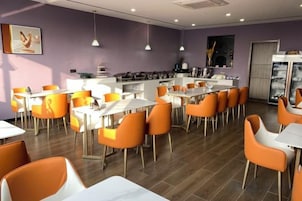 Uma sala de jantar com mesas e cadeiras, uma cozinha ao fundo e uma geladeira.