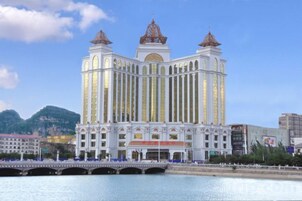 Um edifício grande e ornamentado com uma cúpula central e duas cúpulas menores em cada lado, localizado às margens de um corpo d'água com uma ponte ao primeiro plano.