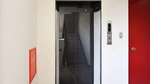 Um corredor com uma porta vermelha, escada e painel de comando de elevador fixado na parede.