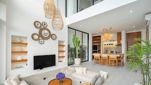 Uma sala de estar moderna com uma televisão de tela plana, uma mesa de centro com um vaso e um sofá. Há uma área de jantar com mesa e cadeiras, e uma cozinha ao fundo.