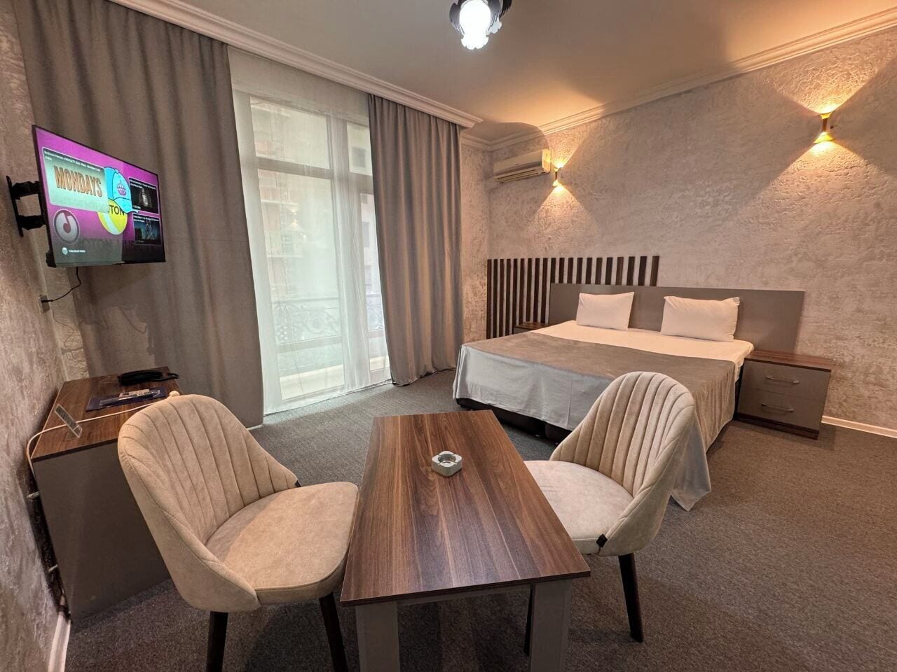 Quarto de hotel com cama, duas poltronas, uma mesinha, televisão e janela com cortinas.