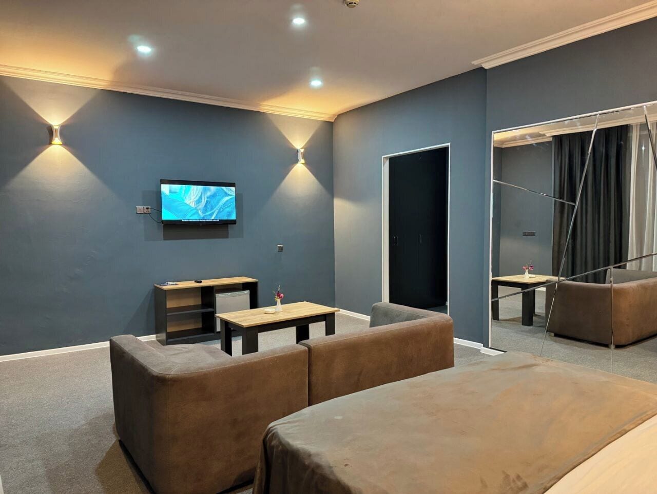 Quarto moderno com televisão de tela plana na parede, sofá, mesa pequena e espelho.
