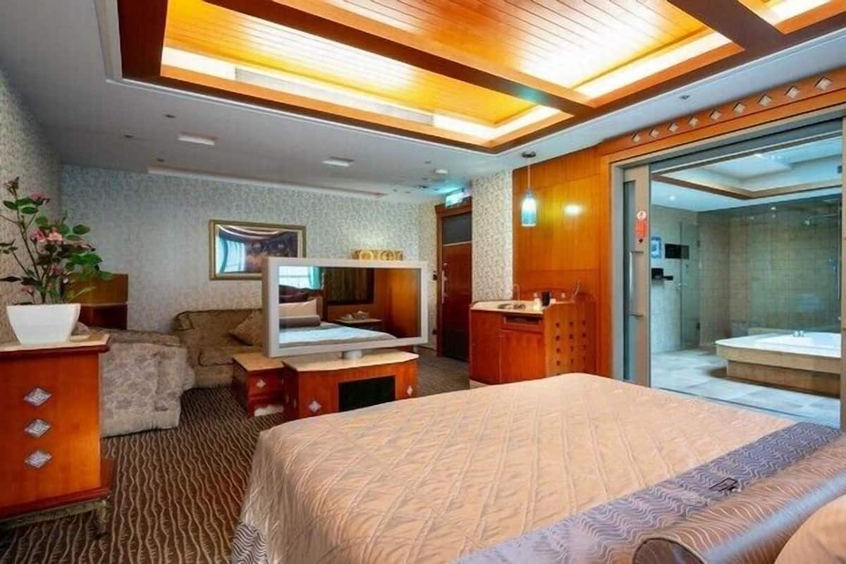 Quarto de hotel com uma cama grande, uma mesa com computador, um sofá, banheiro com chuveiro de vidro e teto de madeira.