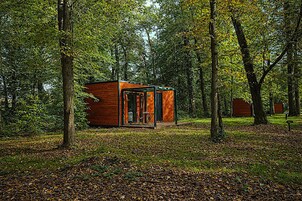 Uma pequena cabana de madeira numa floresta, com uma porta de vidro e uma mesa na parte externa.