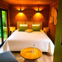 Quarto de madeira com uma cama, duas luminárias fixadas na parede, uma mesa redonda com copos e uma janela com vista.