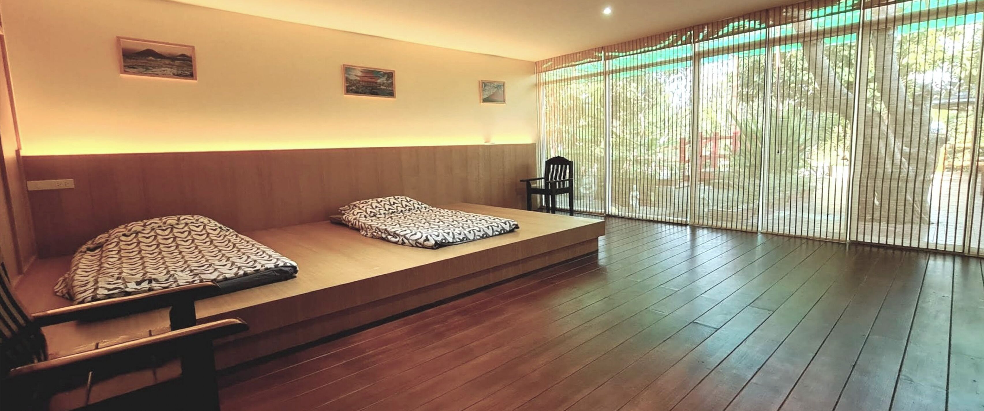 Um quarto com piso de madeira, um sofá com almofadas estampadas, uma cadeira e uma janela grande com persianas verticais.