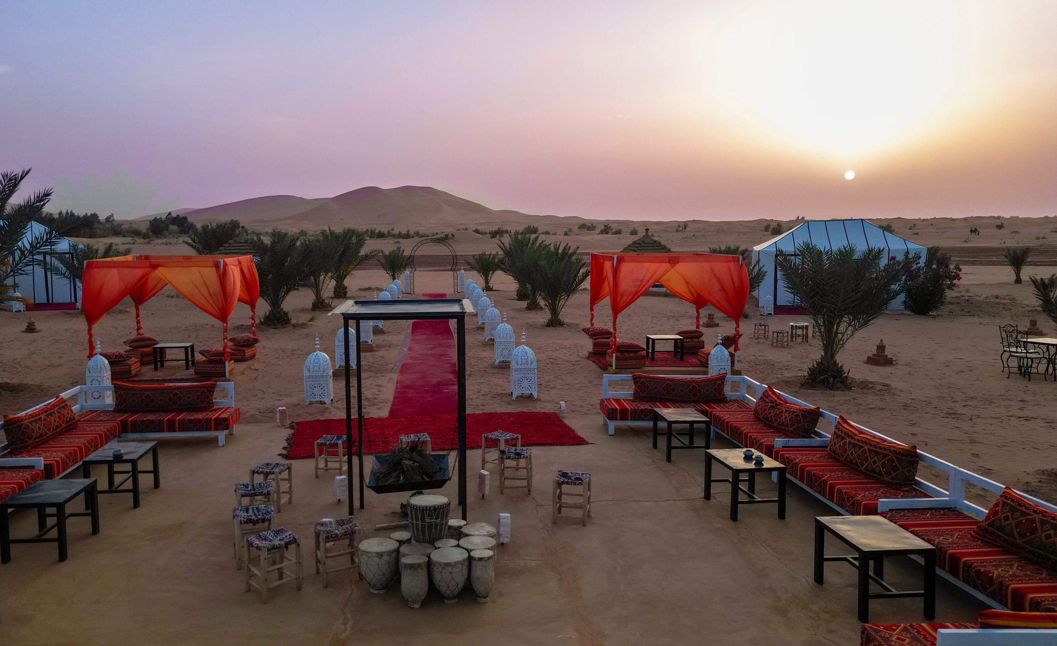 Acampamento no deserto com um tapete vermelho central que leva a uma área de estar com almofadas vermelhas e lanternas brancas.