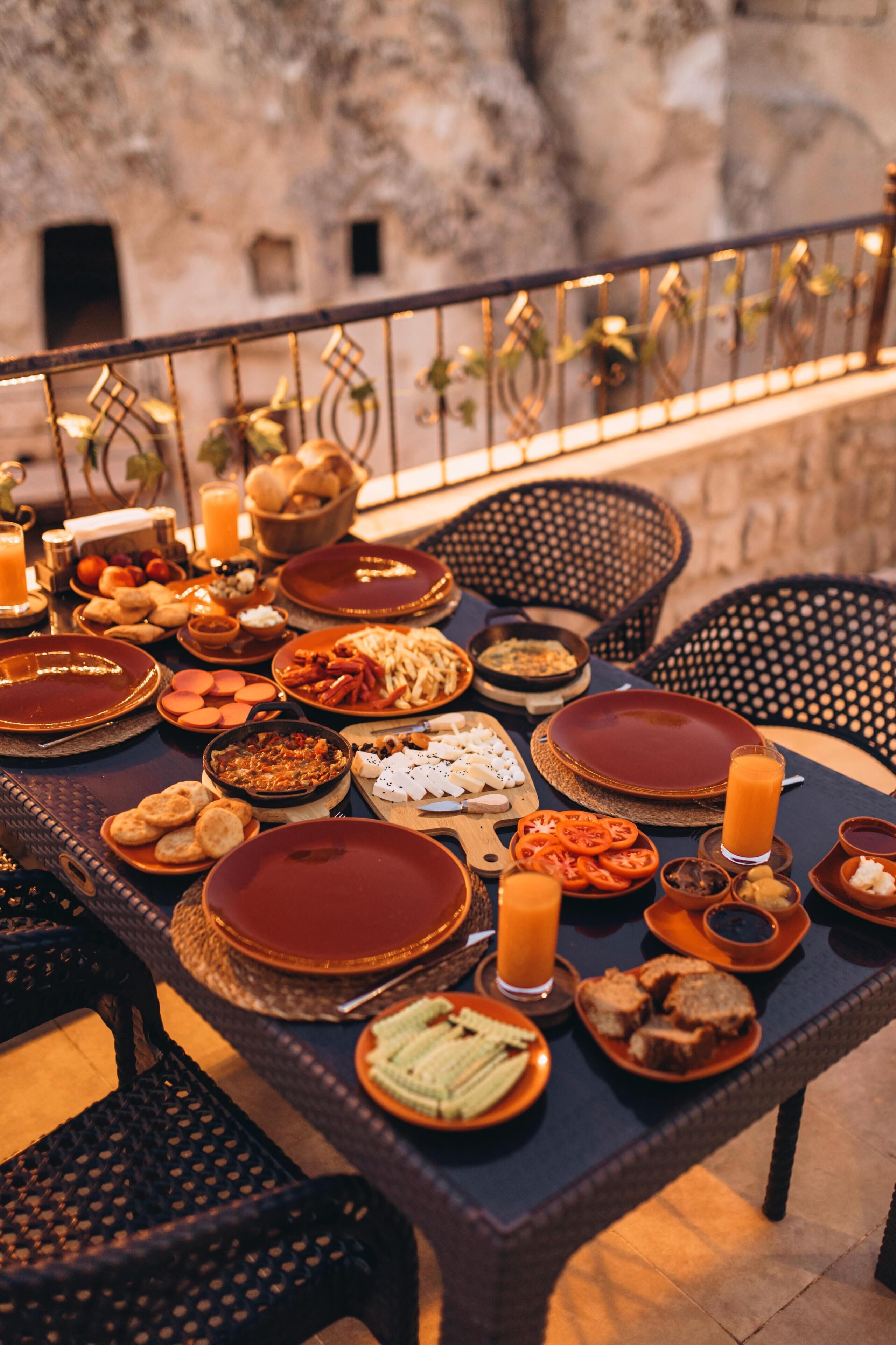 Mesa de jantar posta com diversos pratos e bebidas, cercada por cadeiras de vime, com parede de pedra e guarda-corpo de varanda ao fundo.