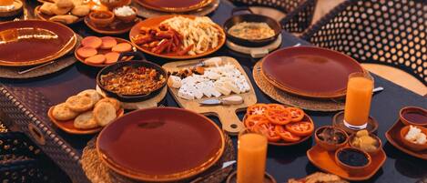 Mesa de jantar posta com diversos pratos e bebidas, cercada por cadeiras de vime, com parede de pedra e guarda-corpo de varanda ao fundo.
