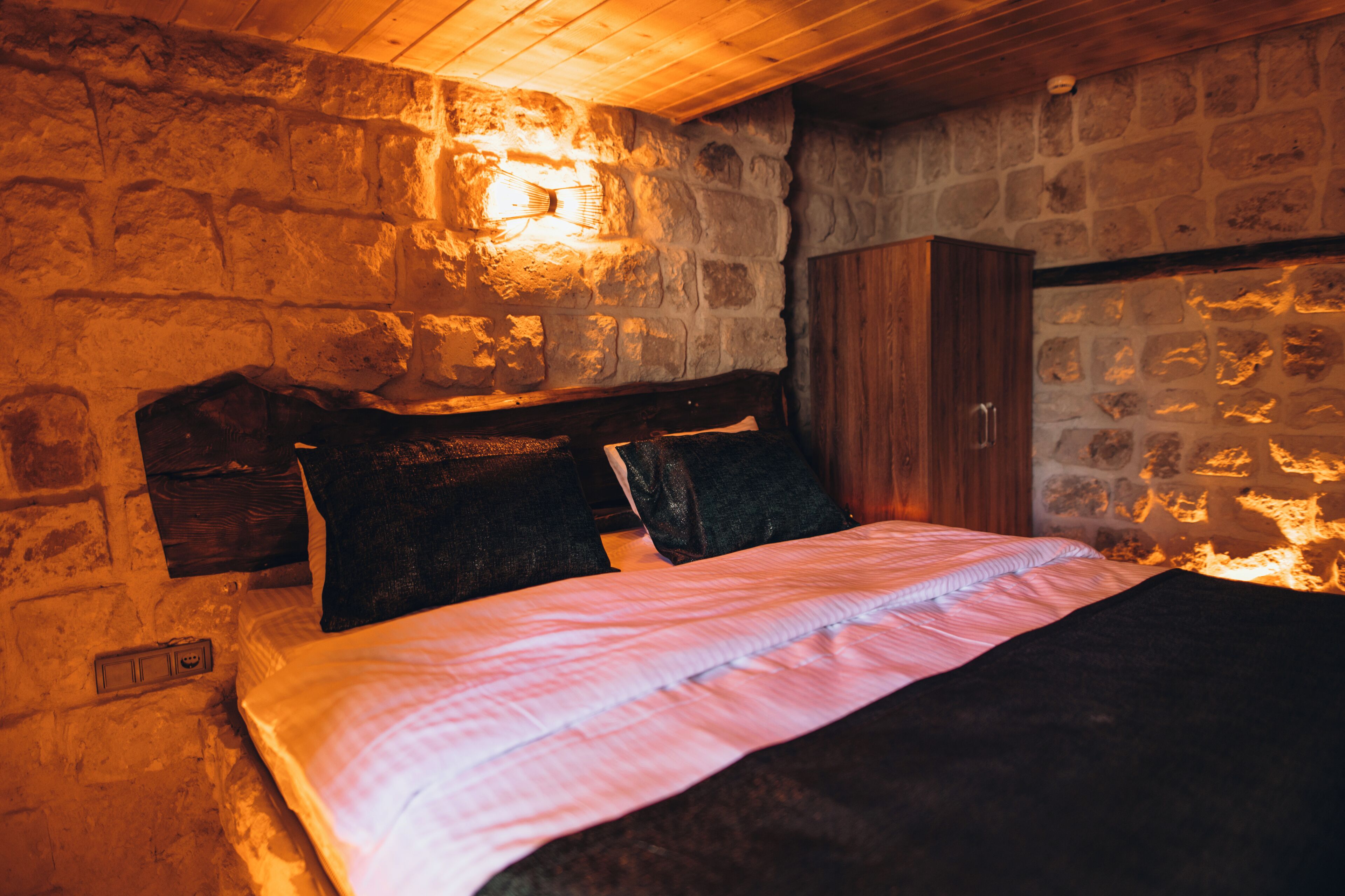Quarto com paredes de pedra, cabeceira de madeira, cama com colcha listrada em rosa e branco, e guarda-roupa de madeira.