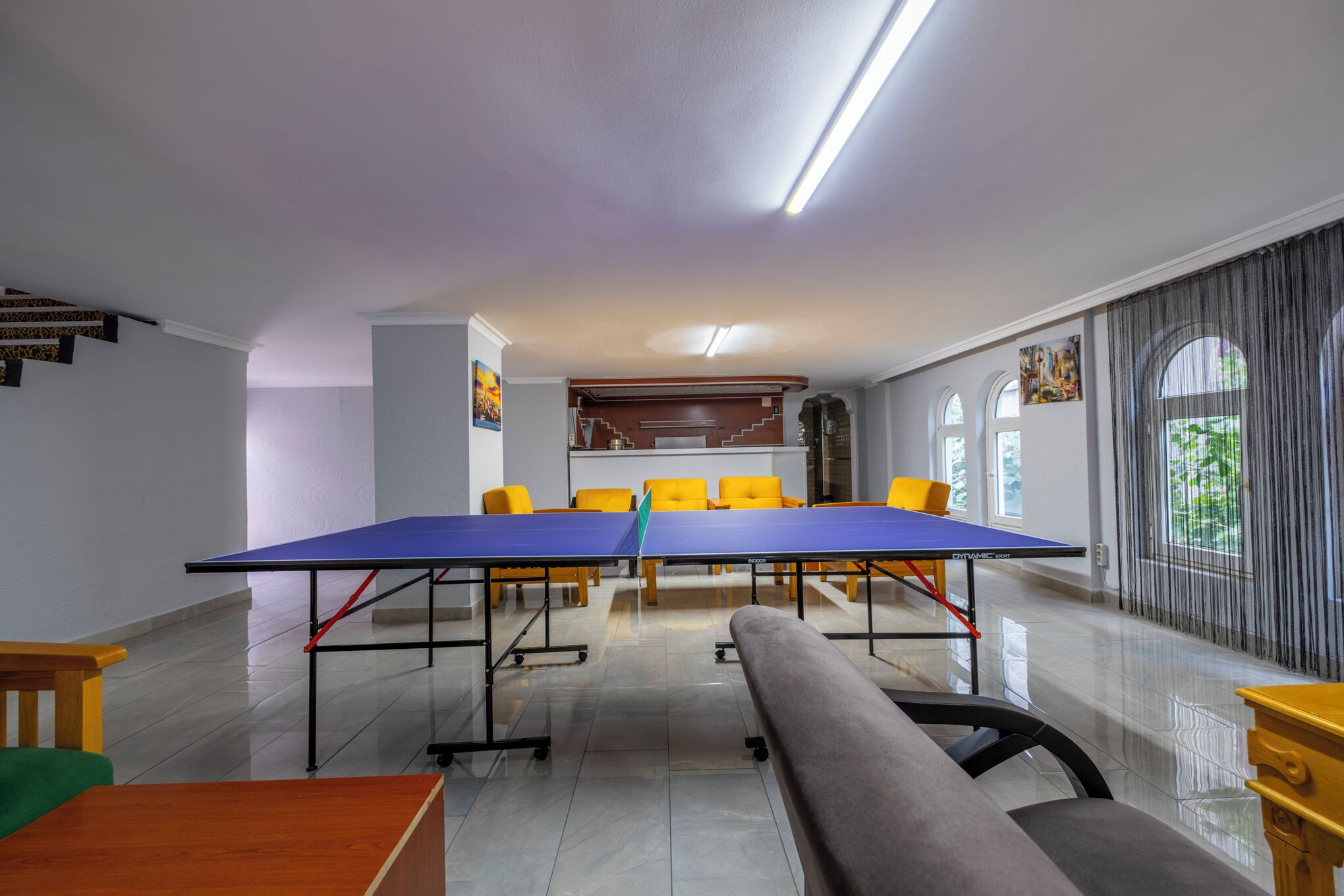 Quarto com mesa de pingue-pongue, cadeiras amarelas, sofá cinza e armário de madeira.