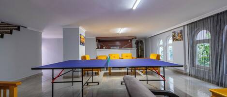 Quarto com mesa de pingue-pongue, cadeiras amarelas, sofá cinza e armário de madeira.