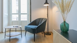 Quarto com cadeira moderna, uma pequena mesa redonda, luminária de chão alta e um vaso verde com plumas de capim-do-campo.