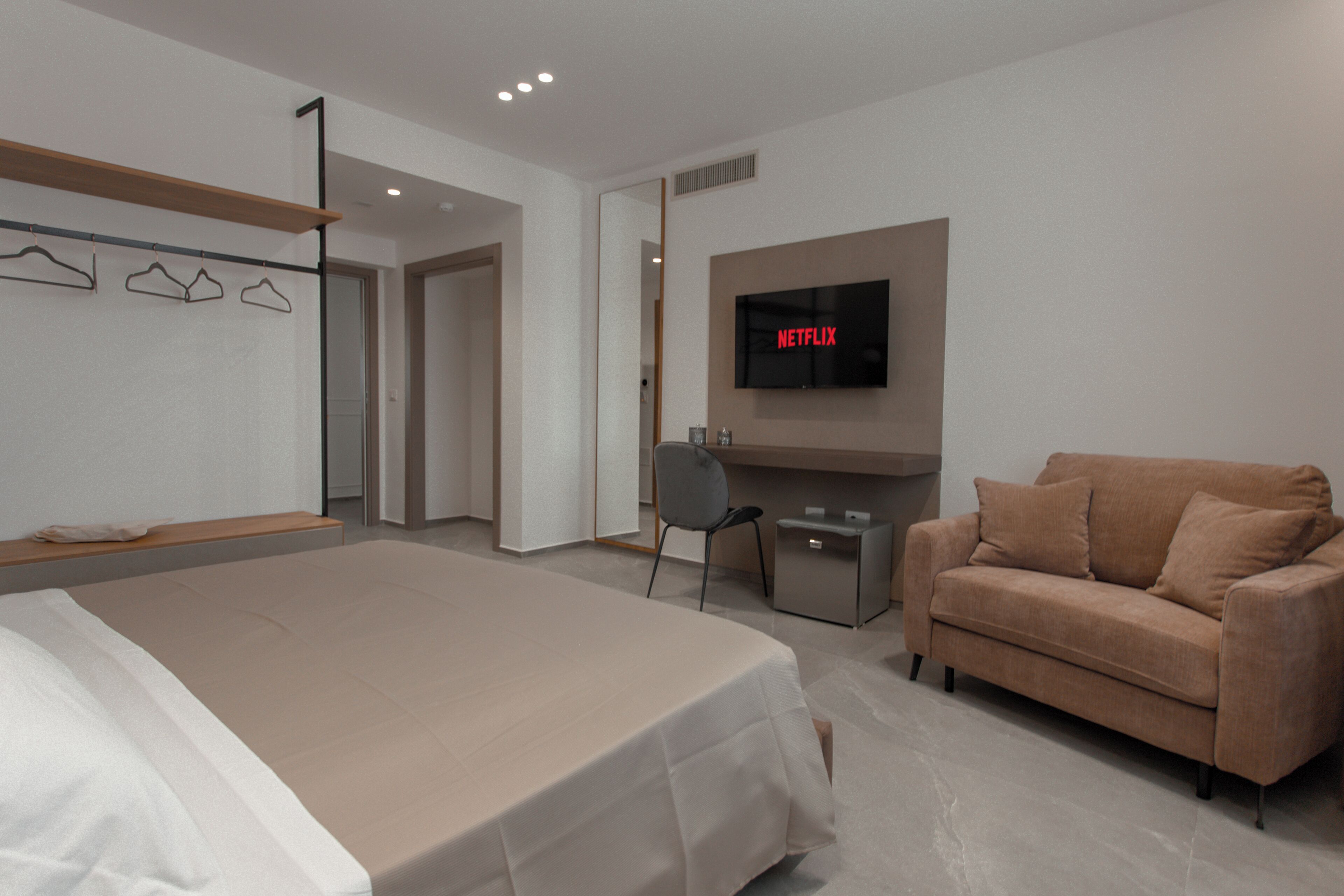 Quarto de hotel moderno com cama grande, sofá, televisão na parede e um cabideiro.