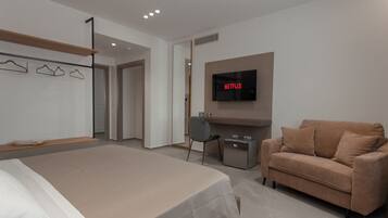 Quarto de hotel moderno com cama grande, sofá, televisão na parede e um cabideiro.