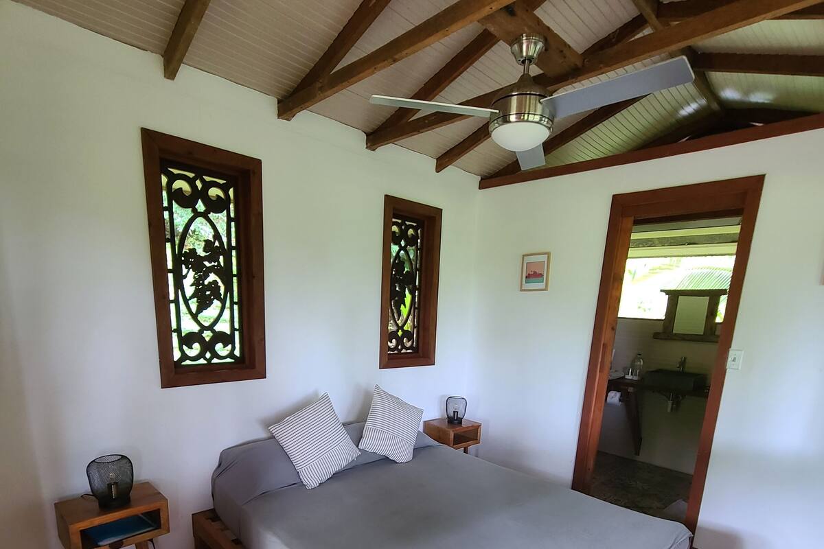 Quarto com cama de madeira, duas mesas de cabeceira, um ventilador de teto e duas janelas decorativas.