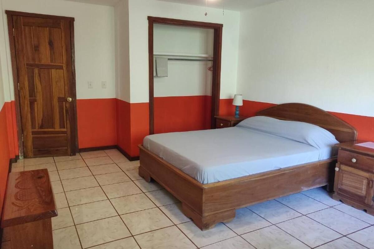 Um quarto com cama de madeira, mesa de cabeceira, banco de madeira e uma porta.