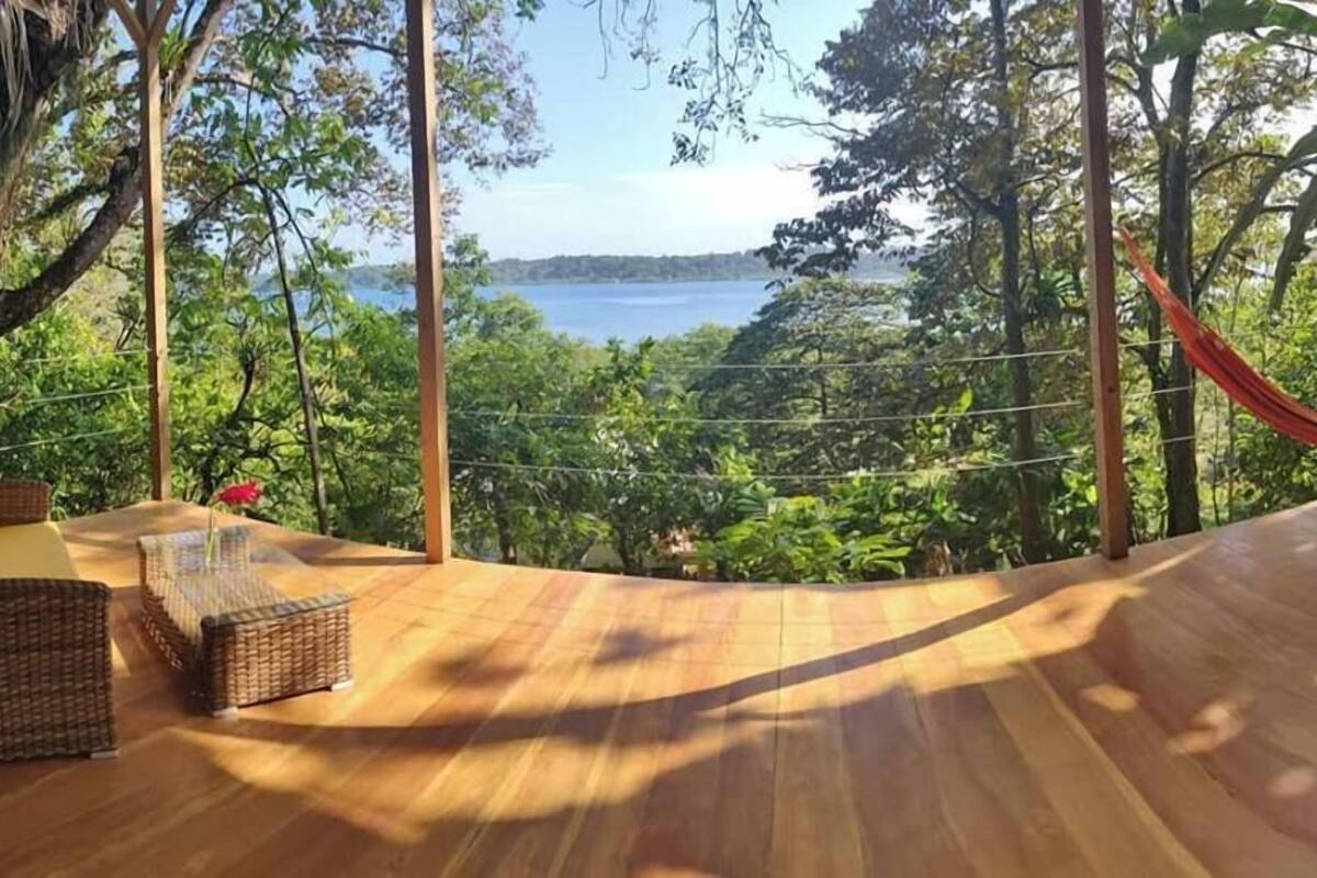 Um deck de madeira com móveis de vime e uma rede, com vista para um lago.