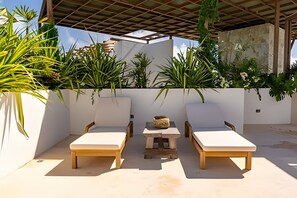 Uma área de estar coberta ao ar livre com duas poltronas e uma pequena mesa, cercada por vegetação exuberante e uma parede branca.