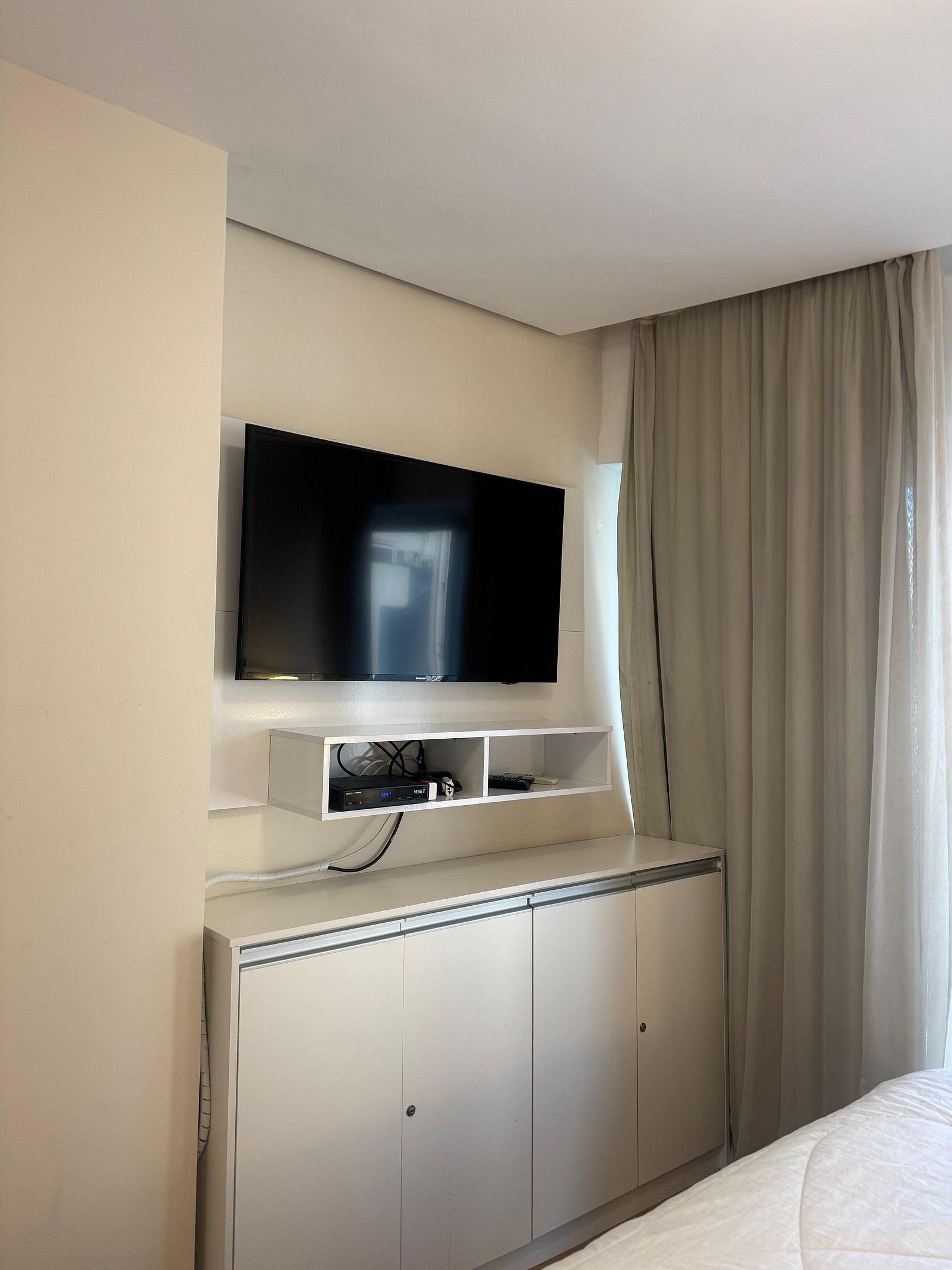 Quarto de hotel com televisão de tela plana na parede, um minibar branco e uma cama com edredom branco.