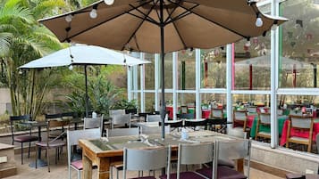 Um café ao ar livre com mesas e cadeiras sob guarda-sóis, cercado por grandes janelas e vegetação.
