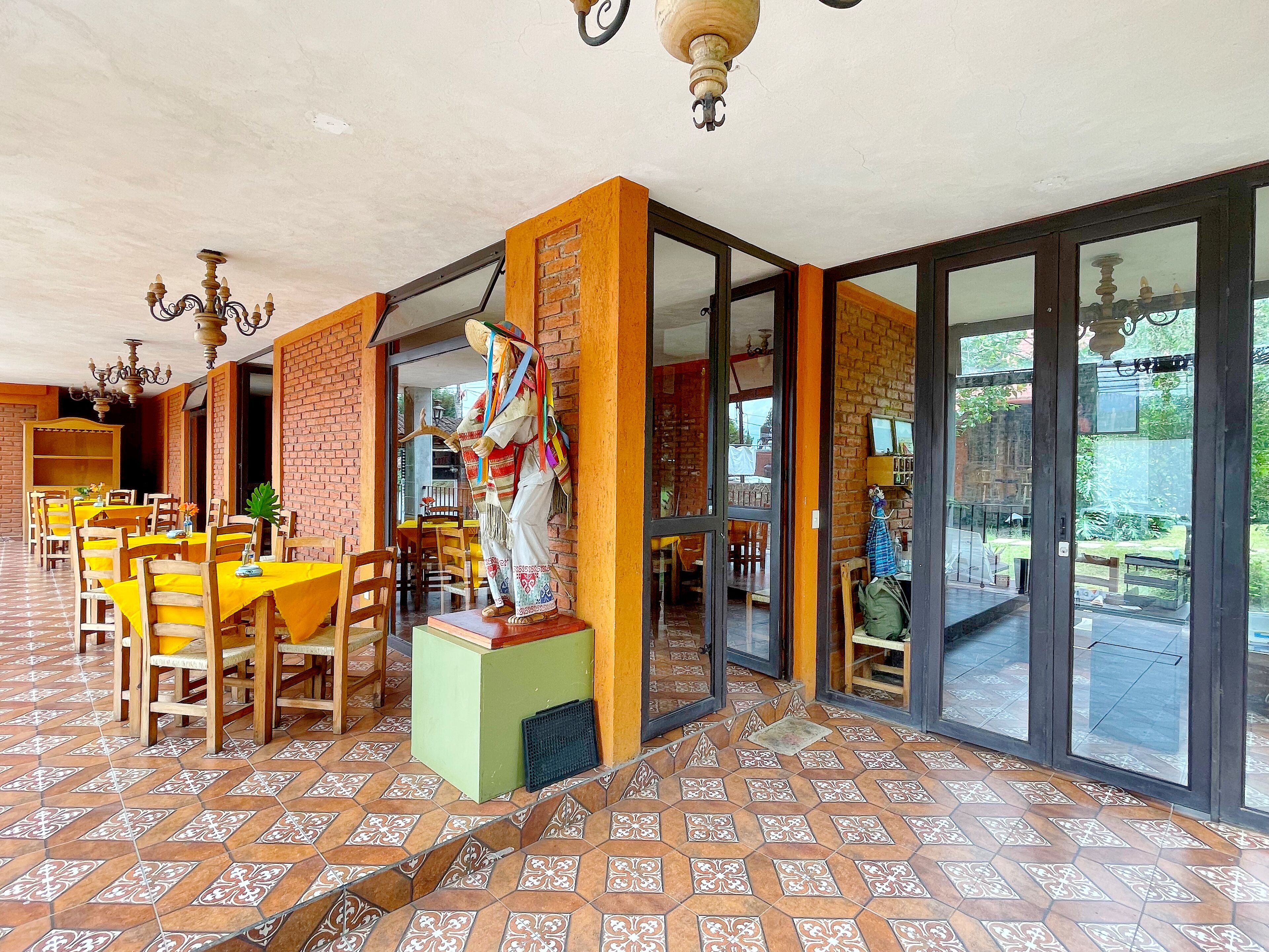 Restaurante com parede de tijolos, grandes janelas e uma estátua de uma pessoa. Mesas montadas com toalhas amarelas e cadeiras de madeira.