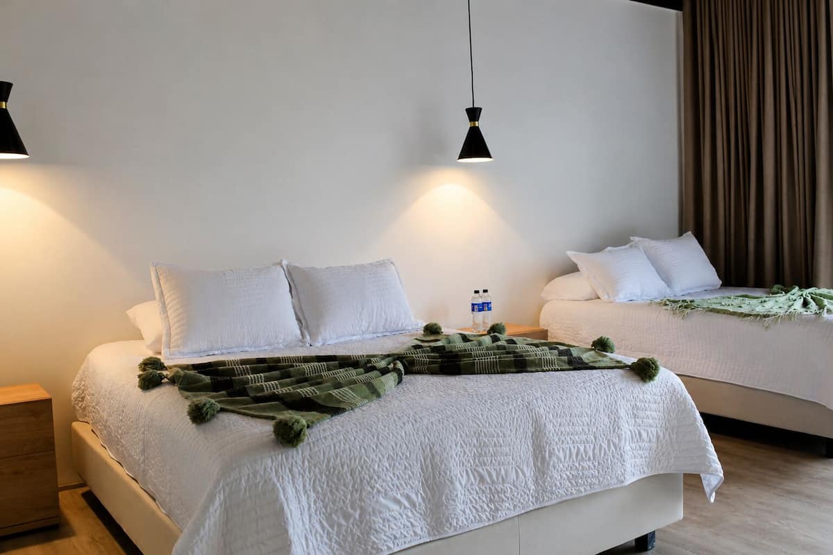 Quarto de hotel com duas camas, cada uma com roupa de cama branca e manta verde. Duas luminárias pendentes pretas estão sobre as camas.