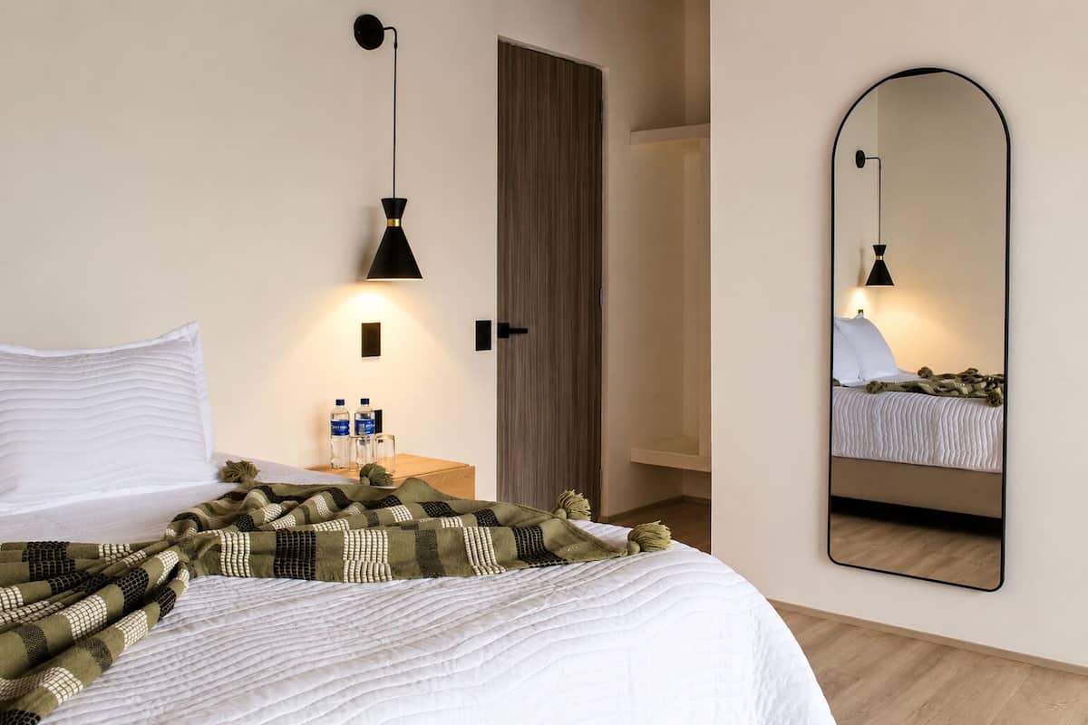 Quarto de hotel com cama, mesa de cabeceira com garrafas de água, espelho e porta.
