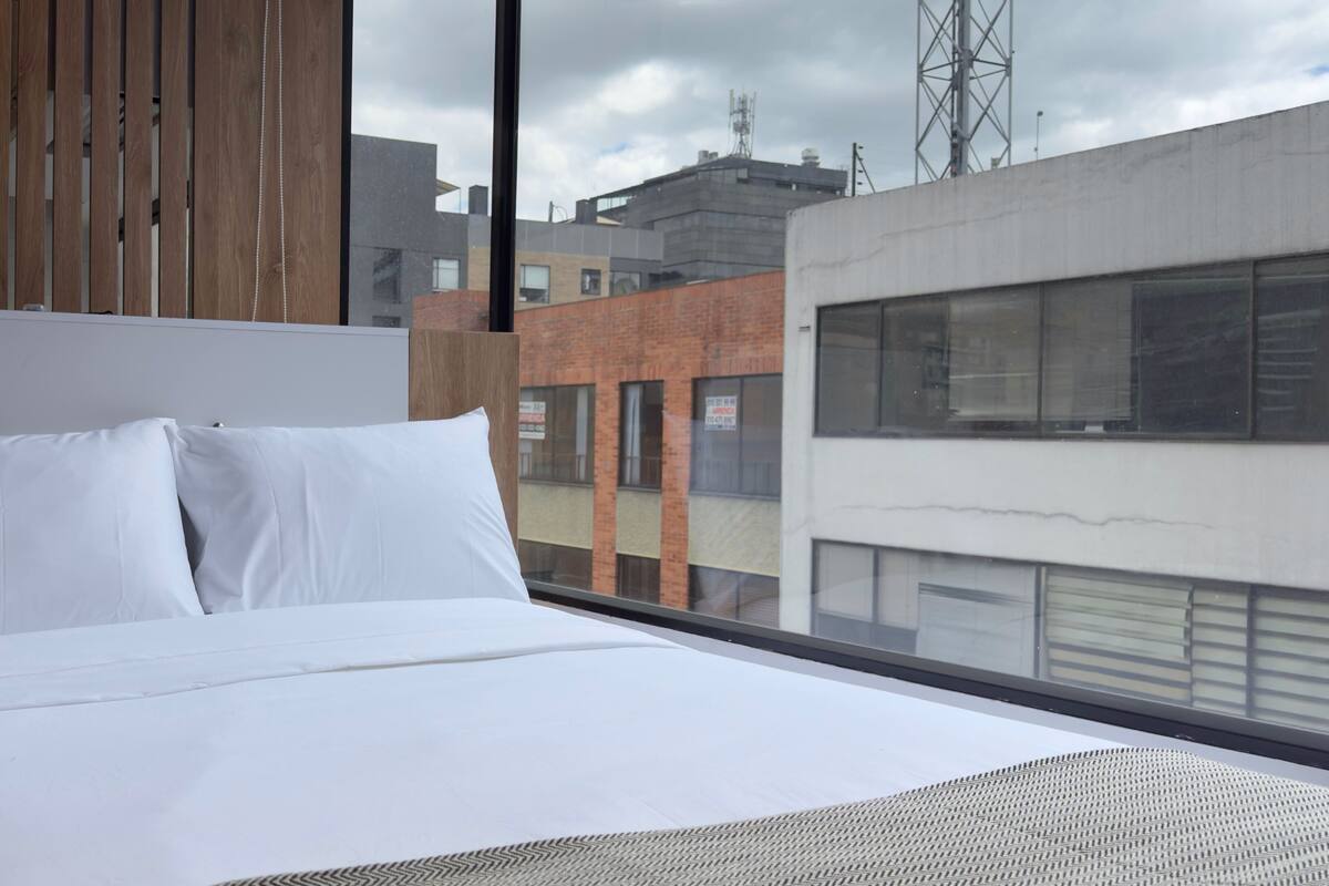 Quarto de hotel com uma cama grande, parede de painel de madeira e vista da cidade através de uma janela ampla.