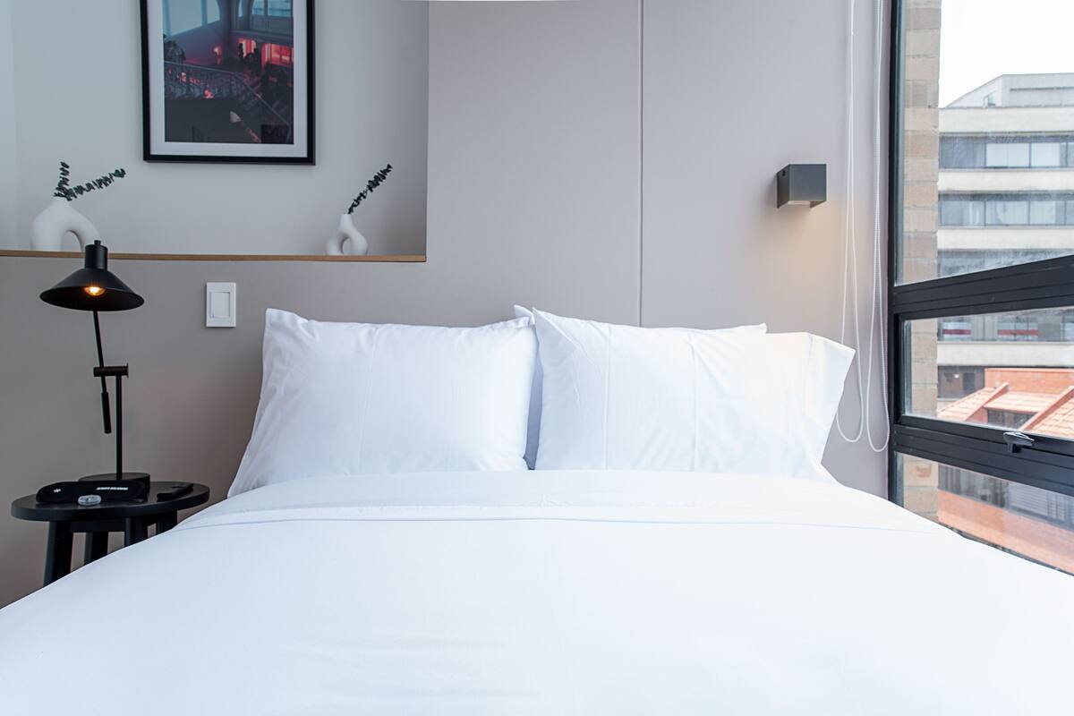 Quarto de hotel com cama, escrivaninha com luminária, quadro na parede e janela com vista para edifícios.