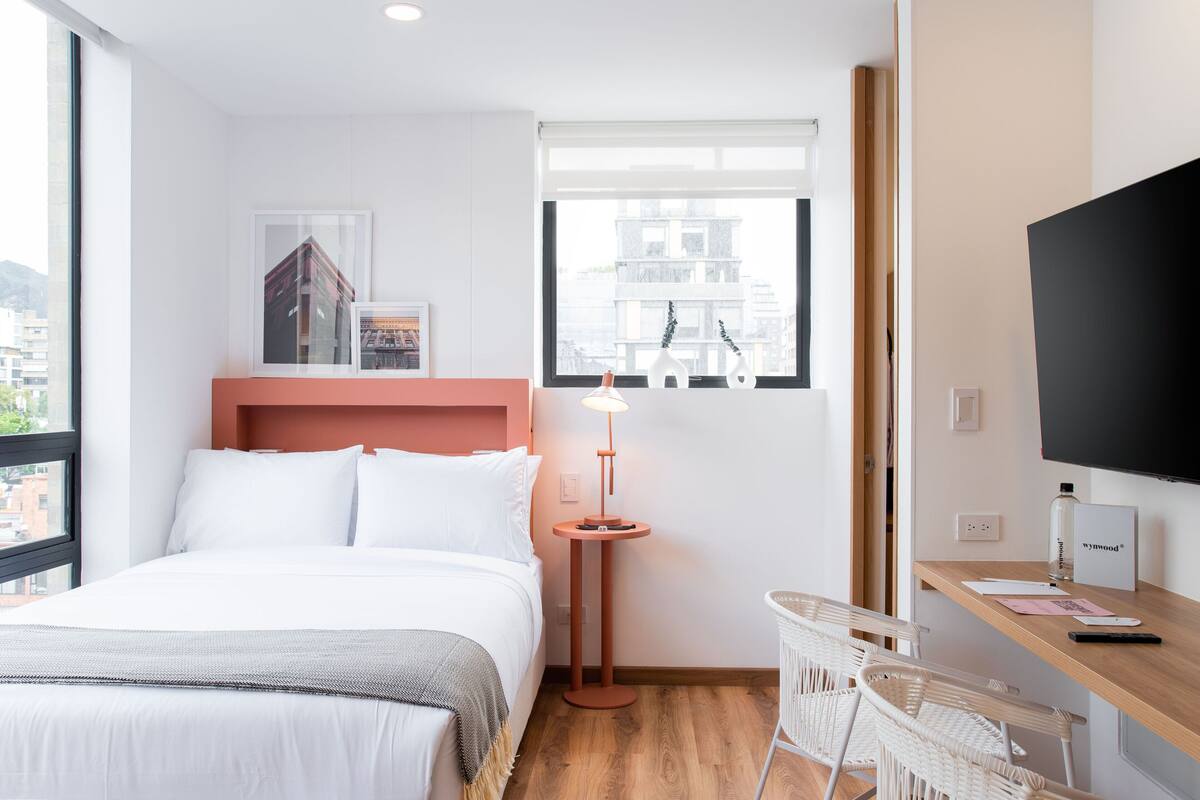 Quarto de hotel com cama, escrivaninha, cadeira, abajur e televisão.