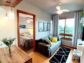 Um quarto aconchegante com sofá, cama com manta xadrez, mesa de madeira com vaso e janela com cortinas.