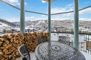 Uma varanda com mesa e cadeiras, uma pilha de lenha e vista para uma paisagem montanhosa coberta de neve.