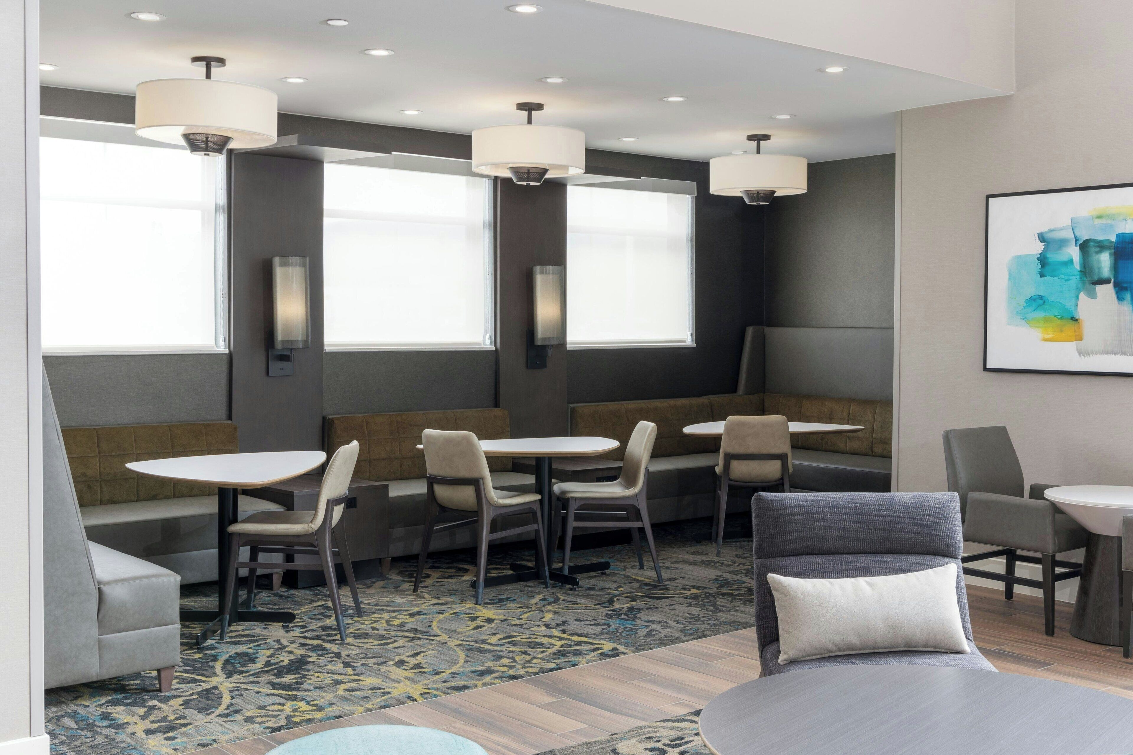 Quarto de hotel moderno com área de estar, mesas redondas e poltronas confortáveis, carpete estampado e uma grande pintura abstrata na parede.