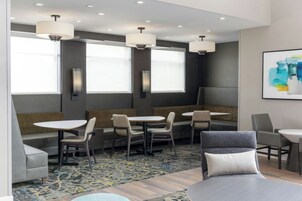 Quarto de hotel moderno com área de estar, mesas redondas e poltronas confortáveis, carpete estampado e uma grande pintura abstrata na parede.
