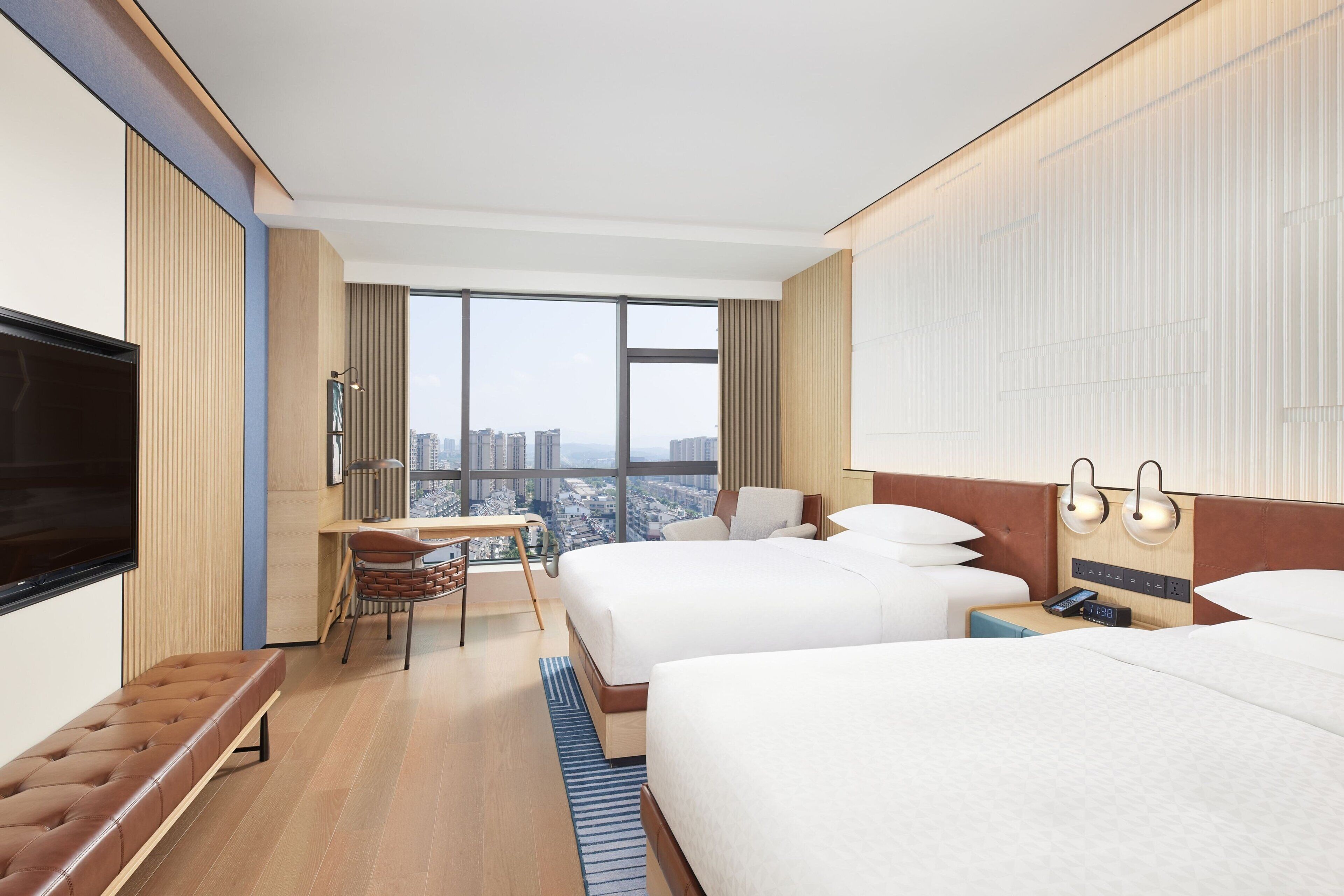 Quarto de hotel com duas camas, uma escrivaninha, uma cadeira, uma televisão e vista para a cidade.