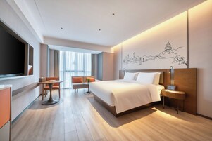 Quarto de hotel moderno com uma cama grande, uma escrivaninha e uma cadeira. Há uma televisão do lado esquerdo do quarto.