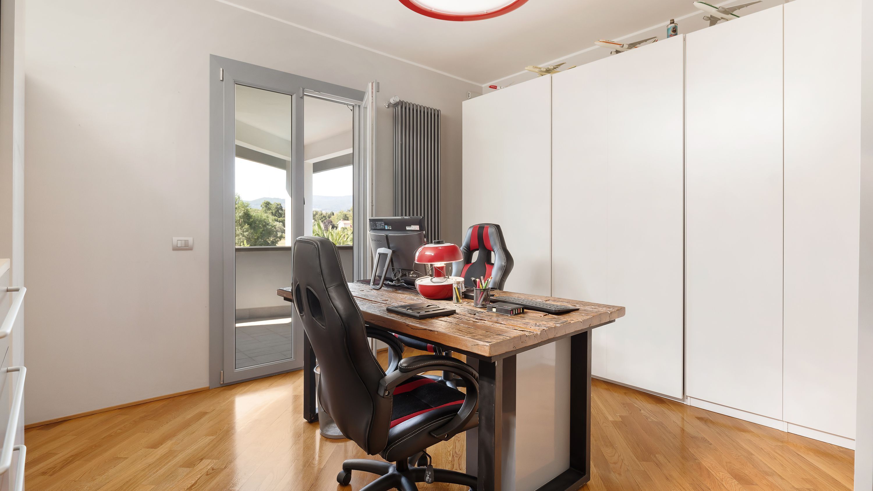 Um espaço de escritório moderno com uma escrivaninha de madeira, uma cadeira de escritório preta, uma luminária de mesa vermelha e um radiador.