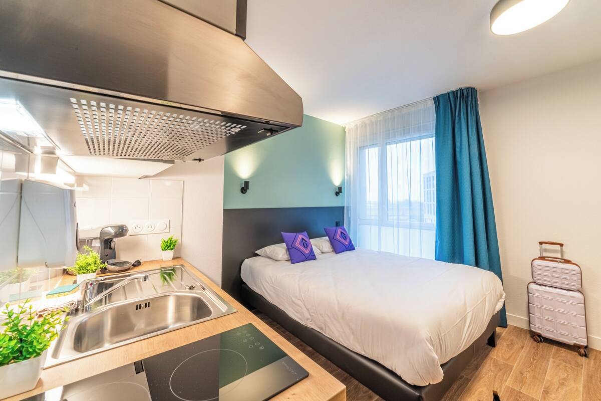 Quarto de hotel moderno com cama, cozinha compacta e mala.