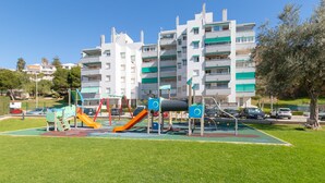 Um parquinho com escorregadores e estruturas de escalada, cercado por uma área gramada e um edifício de vários andares com sacadas.