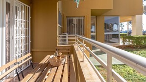 Varanda com um banco de madeira e uma cesta de vime, corrimão branco e vista para a rua com carros estacionados e vegetação.
