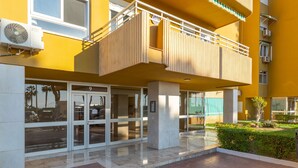 Um edifício com fachada amarela, entrada de vidro e uma pequena área externa com banco e plantas.