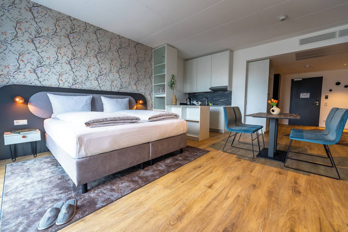 Quarto de hotel moderno com cama, mesa de cabeceira e pequeno espaço de trabalho.