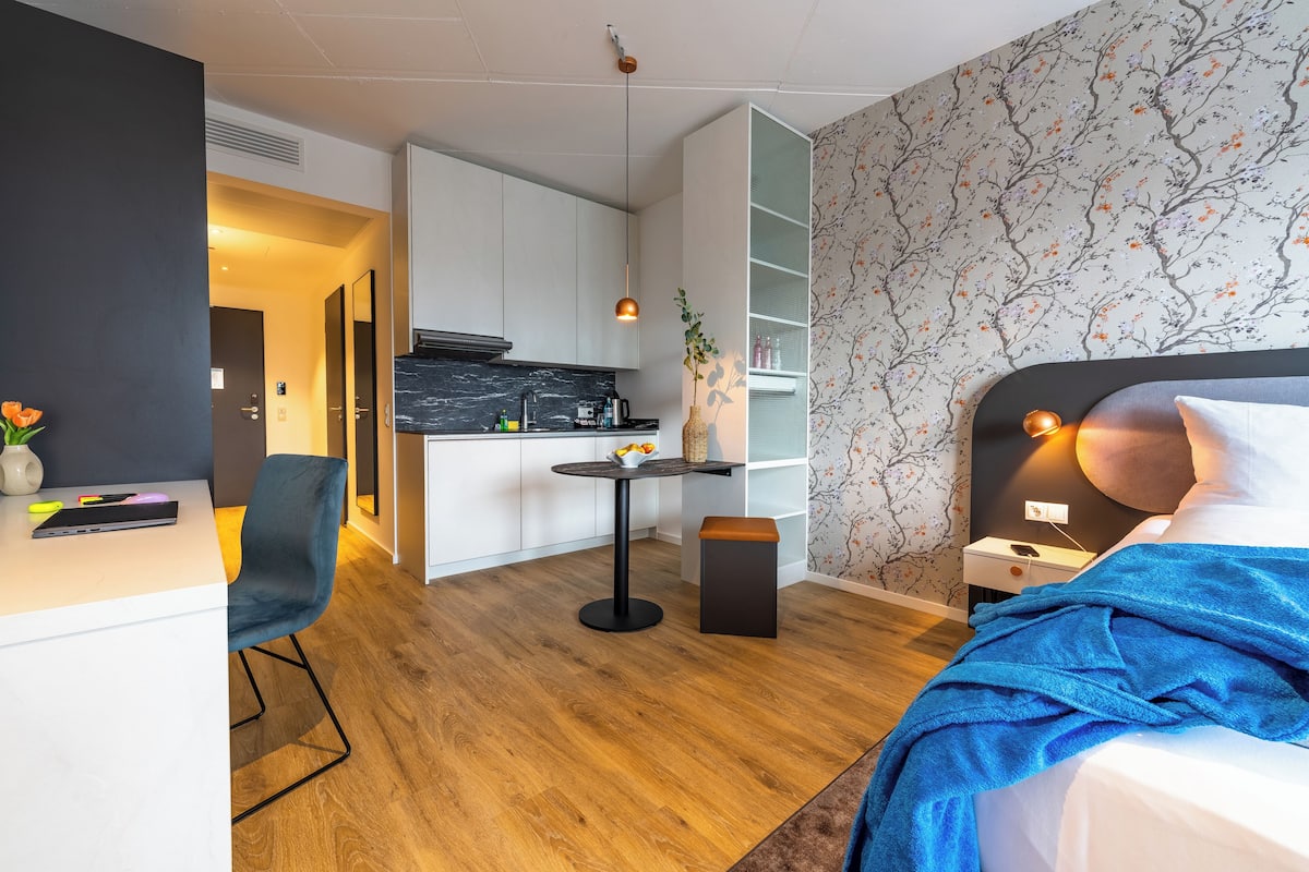 Quarto de hotel moderno com cama, escrivaninha, cadeira, mesa pequena, mesa de cabeceira e cozinha.