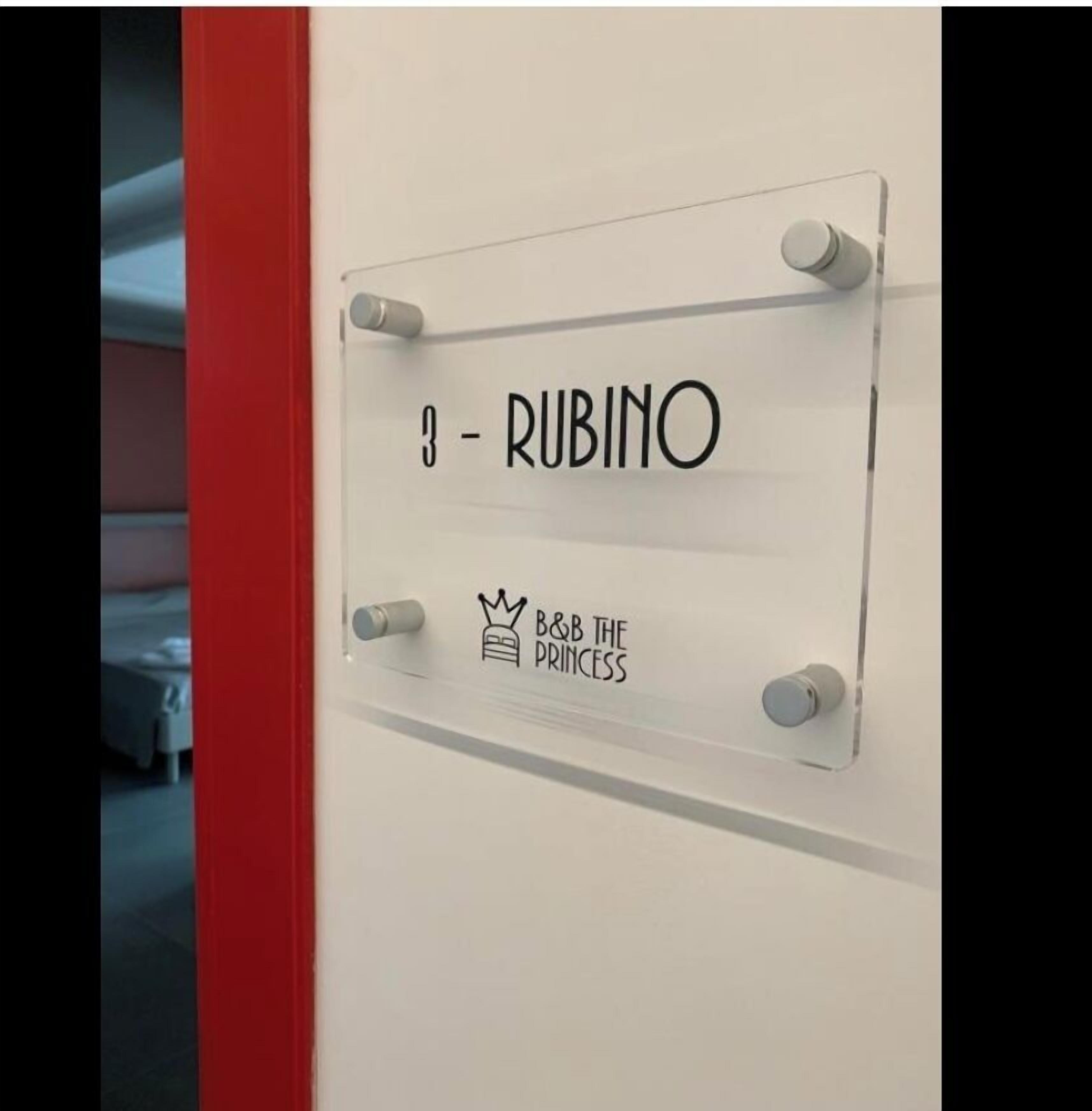 Placa com o número 3 e o nome 'RUBINO' em uma parede branca, com uma pequena coroa e 'BBB THE PRINCESS' abaixo.