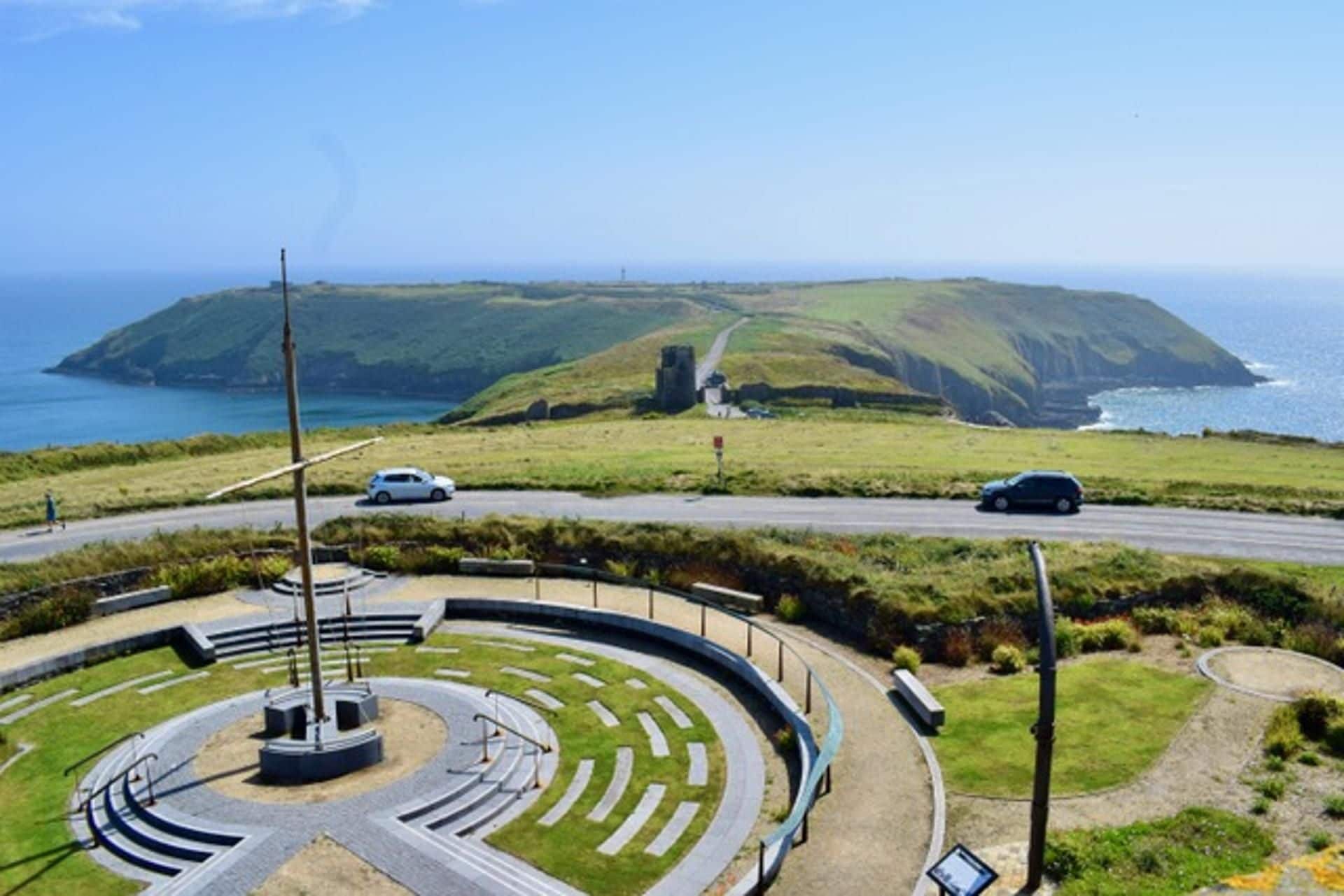 Uma rotatória com um monumento central, uma estrada com carros e uma paisagem costeira ao fundo.