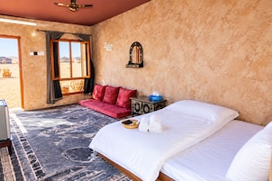 Quarto com uma cama grande, um sofá vermelho, uma mesinha com um estojo de lenços e vista para o deserto por uma porta de vidro.