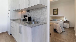 Uma cozinha compacta com bancada de granito, pia embutida e cafeteira. Próximo à cozinha, um quarto com cama, mesa de cabeceira e um quadro na parede.