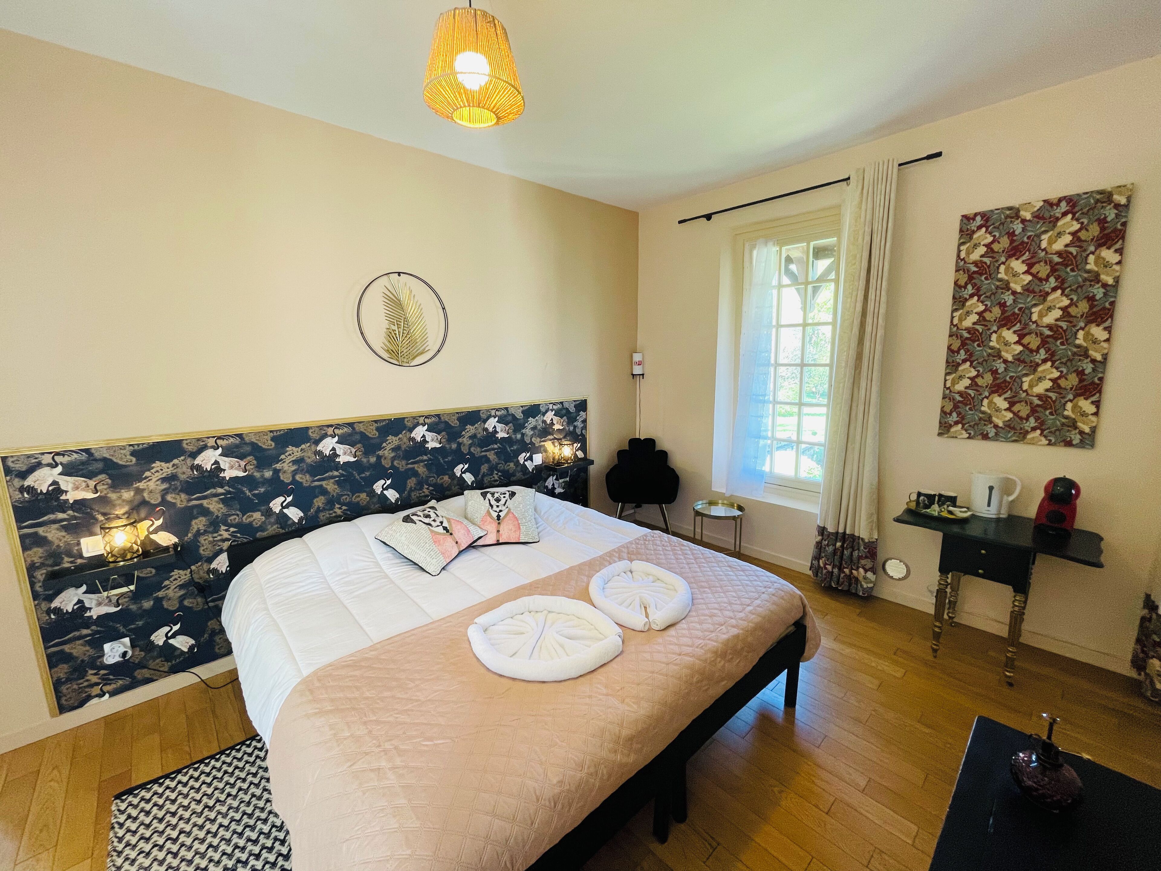 Quarto de hotel com uma cama grande, uma mesa de cabeceira com abajur, uma janela com cortinas e um quadro floral na parede.